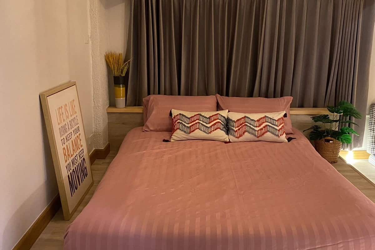 Quarto com uma cama grande, luminária pendente, aparelho de ar condicionado, quadro com frase motivacional e planta em vaso.