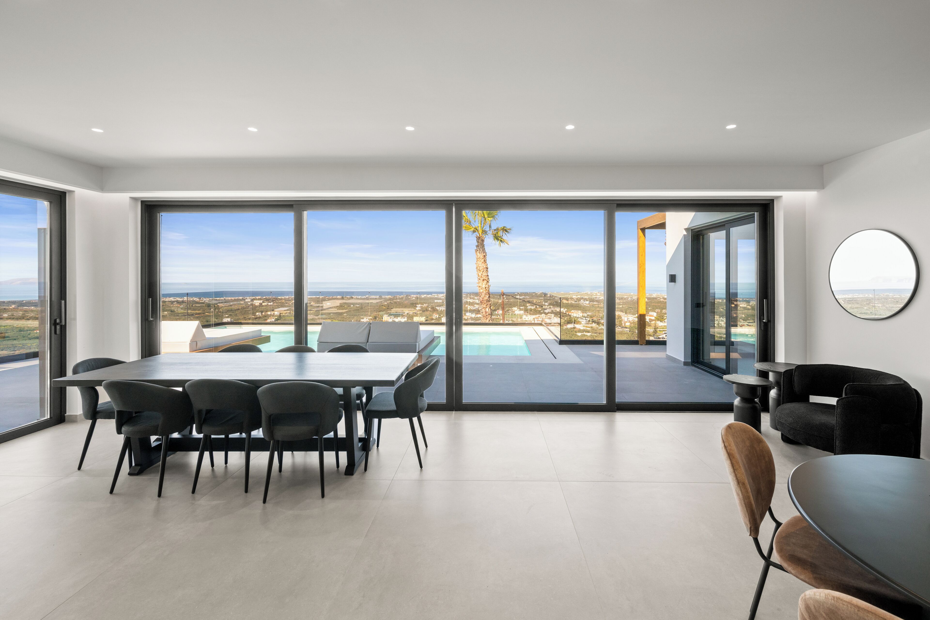 Uma área de jantar moderna com uma mesa grande e cadeiras, janelas amplas com vista para uma piscina e uma palmeira, e um espelho redondo na parede.