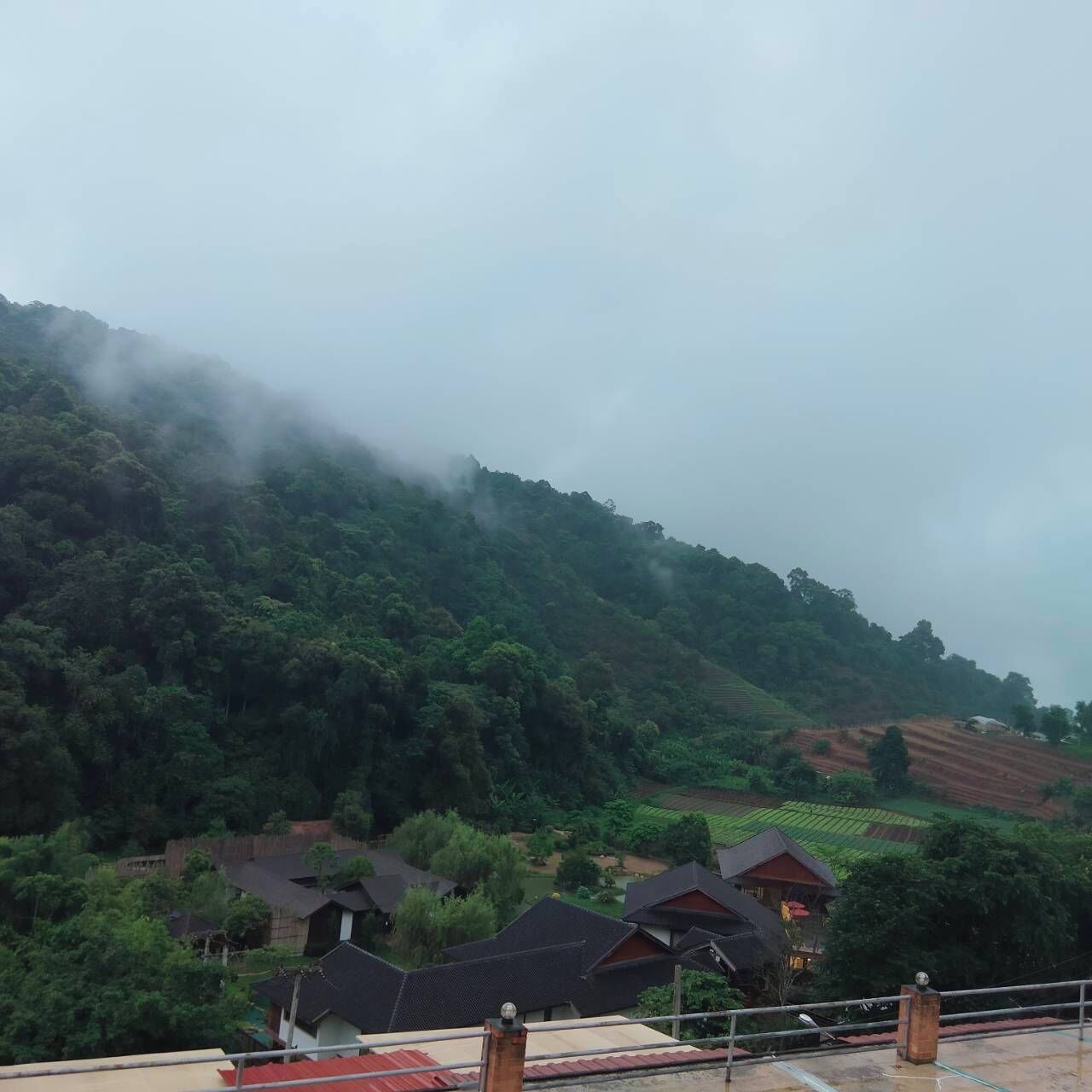 Uma aldeia rural com casas tradicionais, campos em socalcos e uma colina envolta em névoa.