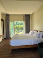 Quarto de hotel com uma cama grande, cabeceira de madeira, poltrona verde e varanda com vista para vegetação.