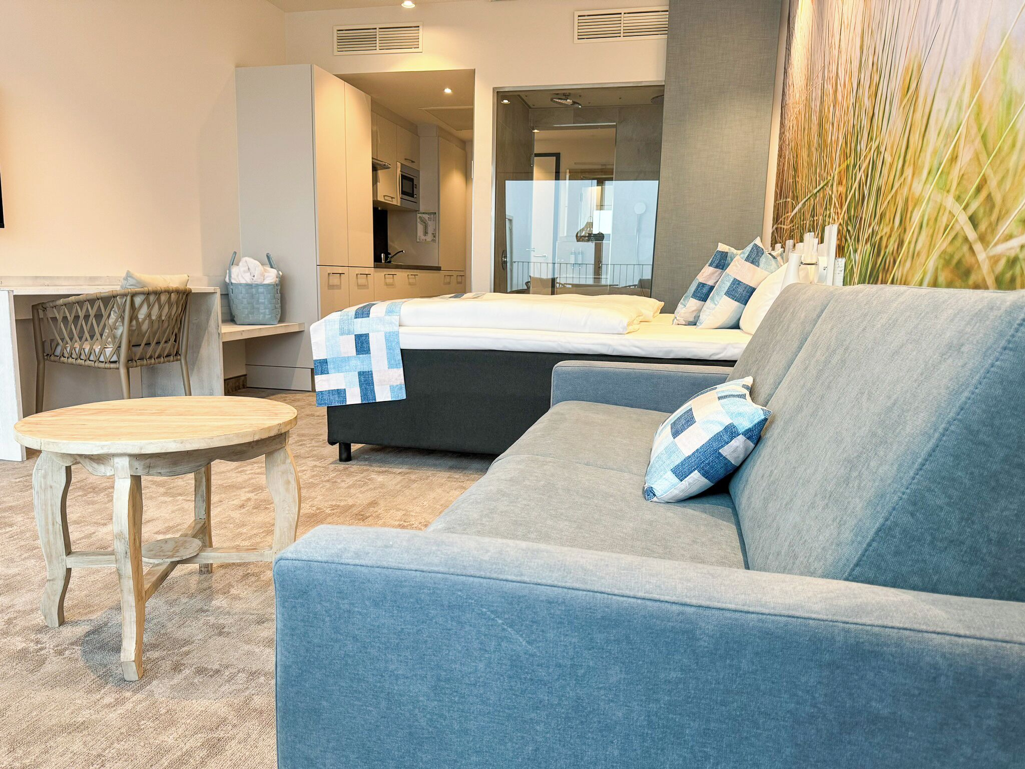 Quarto de hotel moderno com um sofá cinza, uma mesa de madeira e uma cama com cobertor azul e branco xadrez.
