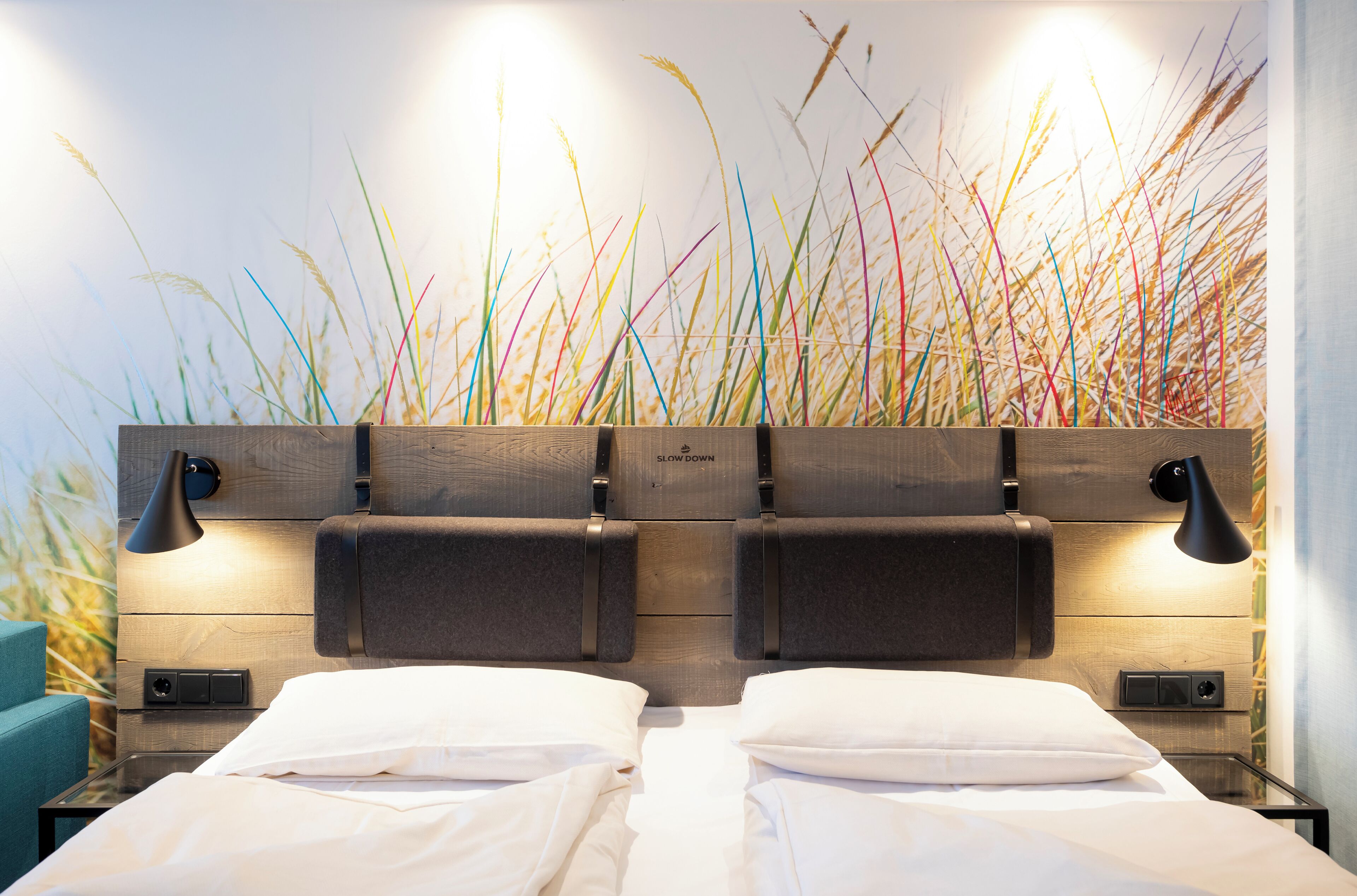 Uma cama de casal com cabeceira que apresenta uma placa com a inscrição 'SLOW DOWN', duas mesas de cabeceira e um abajur fixado na parede.