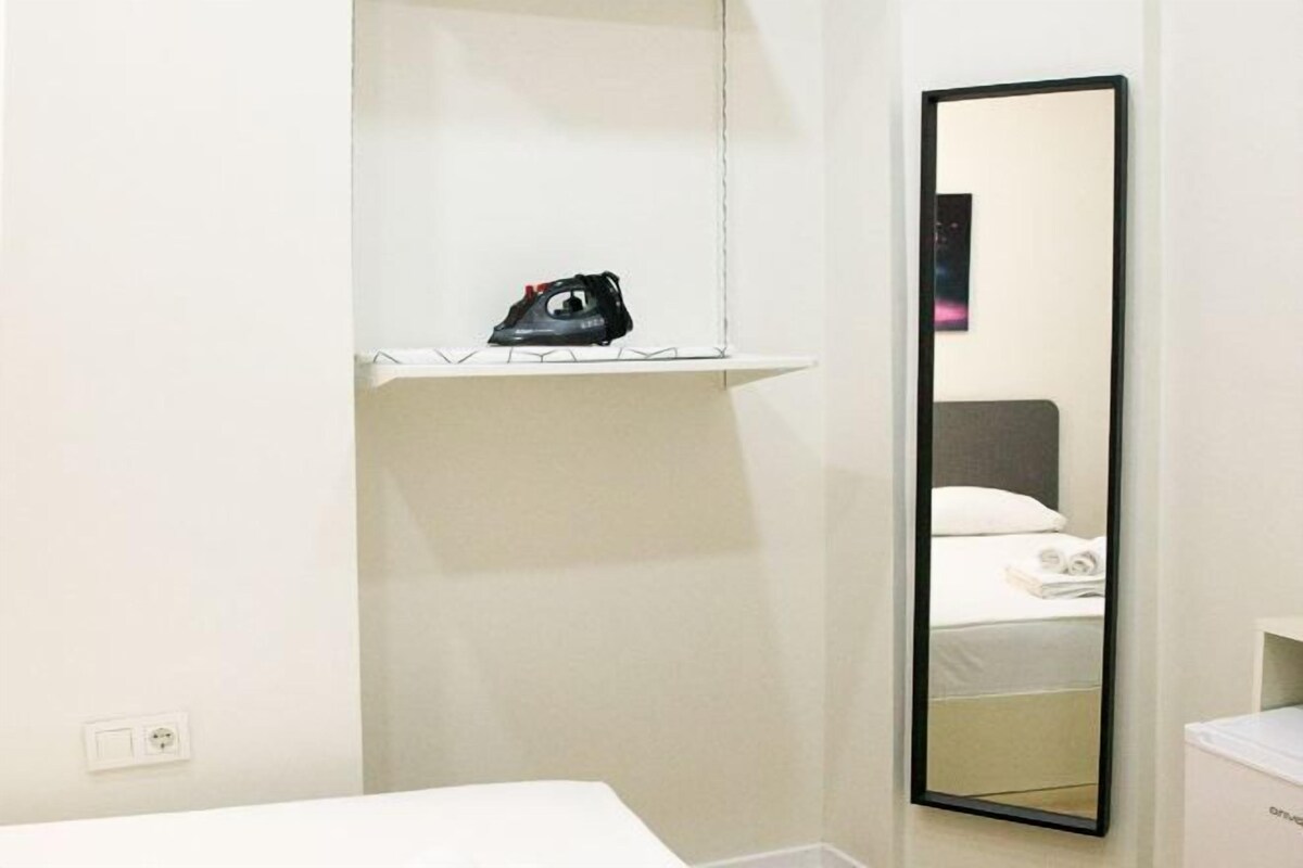 Quarto de hotel com cama, espelho, prateleira com câmera e escrivaninha branca.