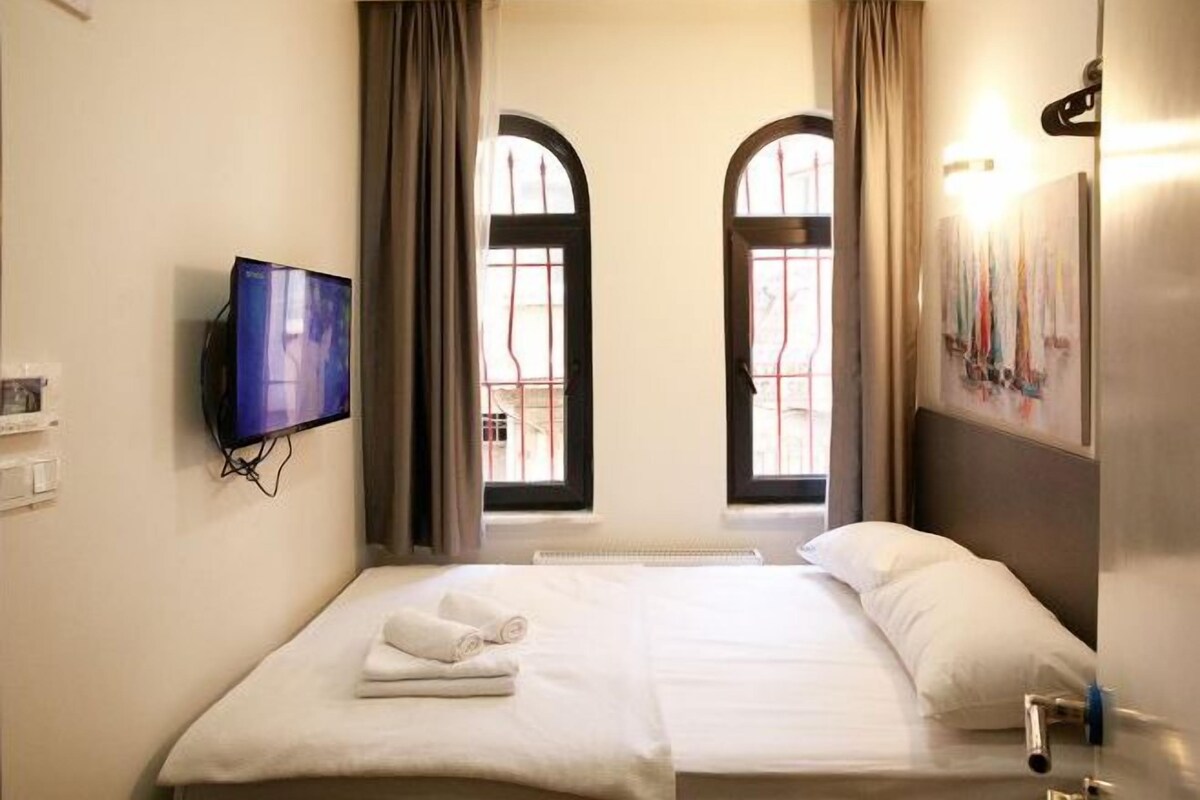 Quarto de hotel com uma cama, uma televisão de tela plana na parede e duas janelas com cortinas.