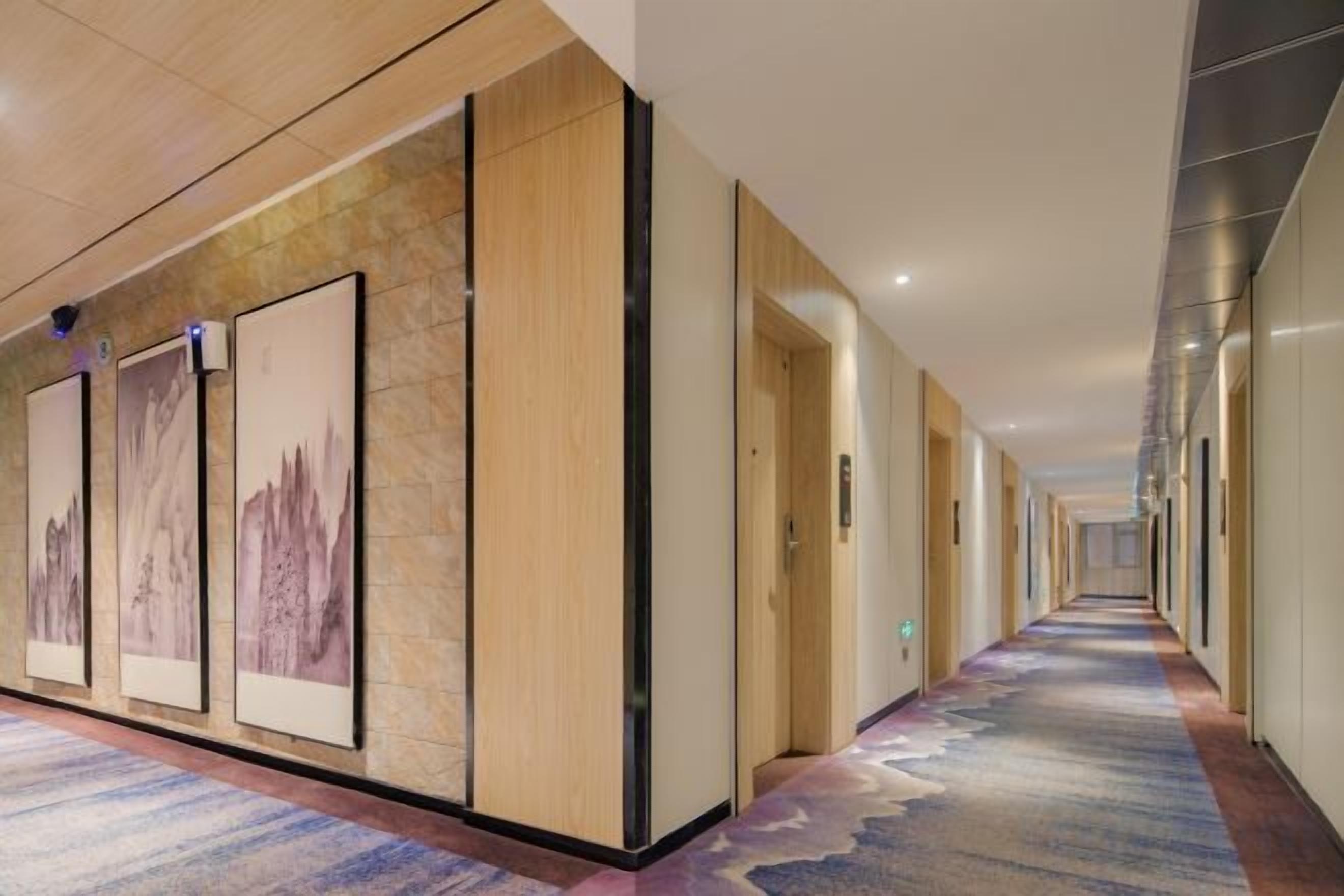 Corredor de hotel com obras de arte emolduradas nas paredes, piso coberto por carpete e portas que dão acesso aos quartos.