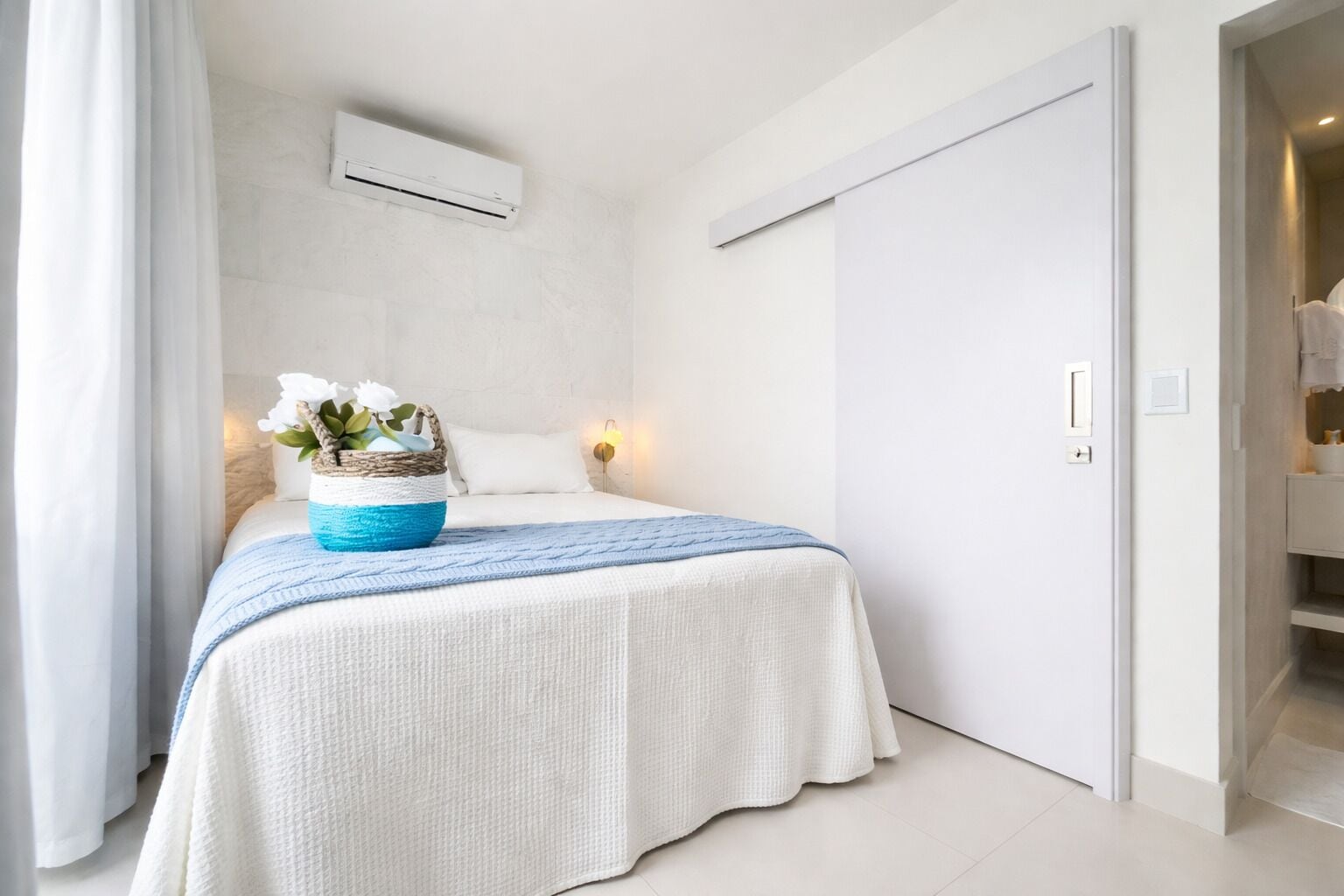 Quarto moderno com cama, ar-condicionado branco, abajur e porta para banheiro.