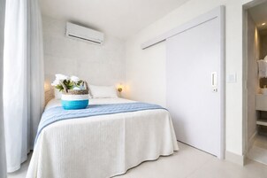 Quarto moderno com cama, ar-condicionado branco, abajur e porta para banheiro.