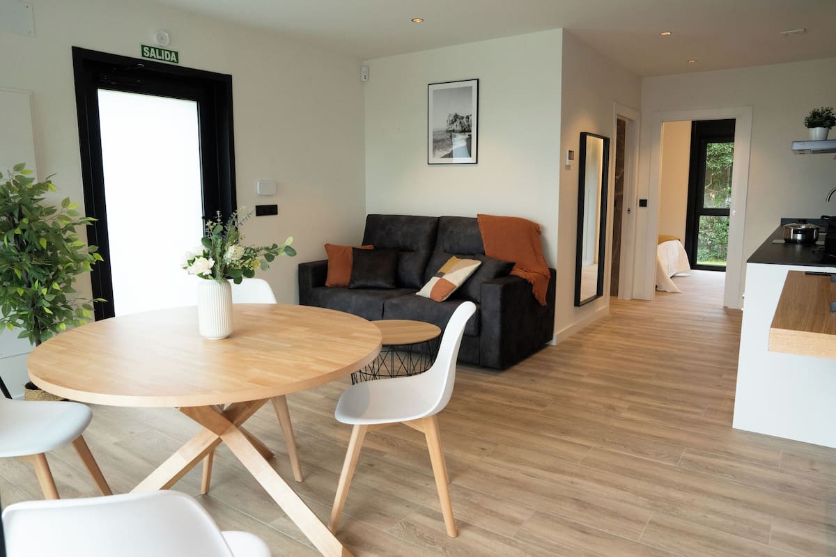 Uma sala de estar moderna com uma mesa de madeira redonda, cadeiras brancas, um sofá cinza com almofadas laranja e um quadro na parede.