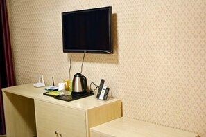 Quarto de hotel com televisão na parede, uma mesa com chaleira, telefone e uma chávena.