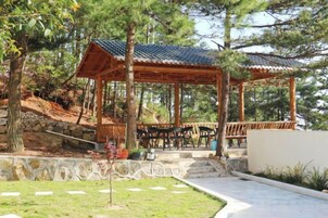 Um gazebo de madeira com telhado de cerâmica, rodeado por muros de pedra e uma escadaria de pedra que leva até ele.