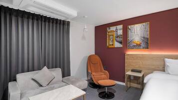 Quarto de hotel com sofá, poltrona, mesa de cabeceira e cama. Há duas pinturas na parede e uma luminária no teto.