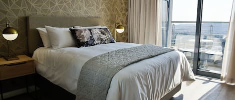 Quarto de hotel moderno com uma cama grande, duas luminárias de cabeceira, parede com padrão geométrico e varanda com vista para a cidade.