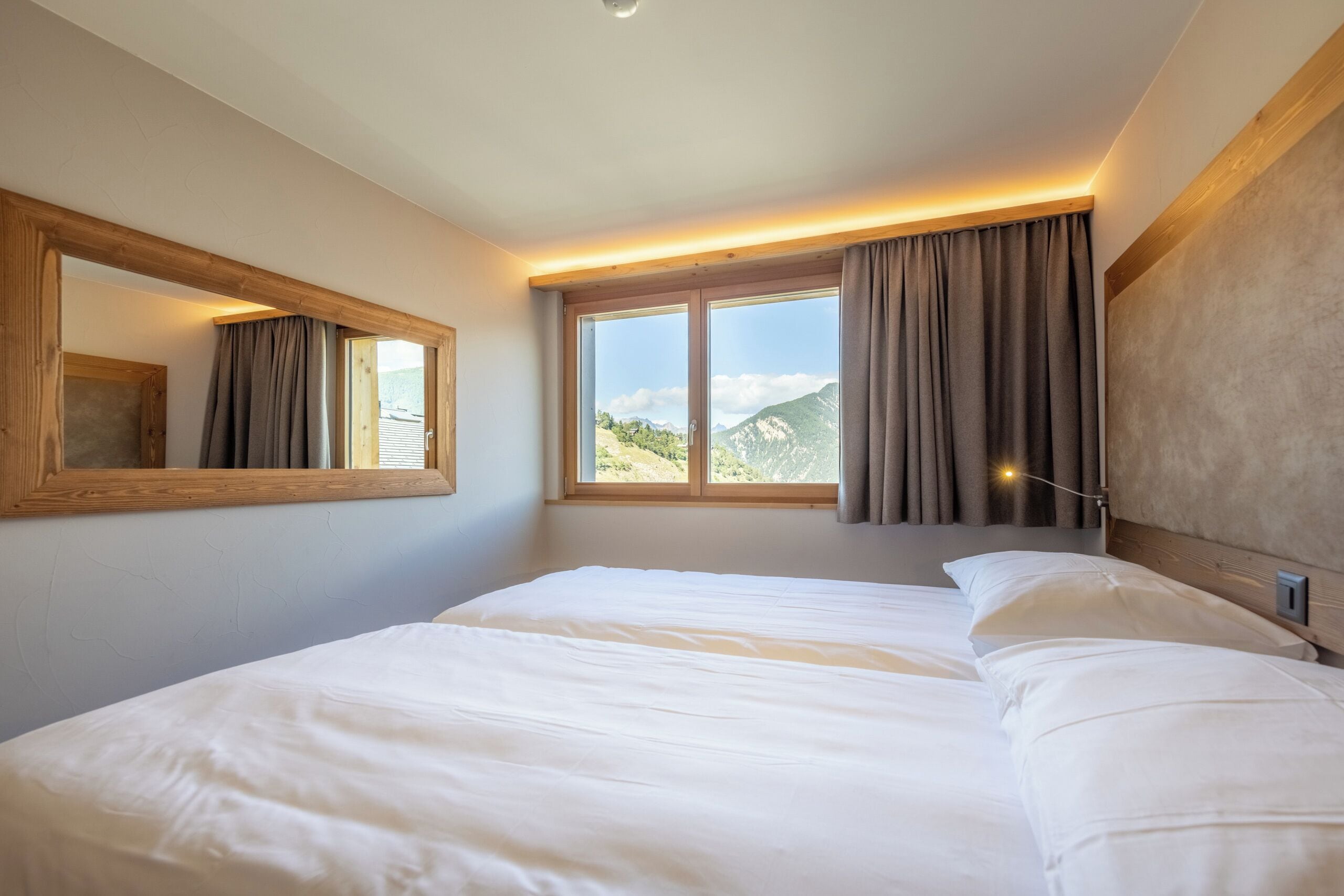 Quarto com cama grande, janela com cortinas, espelho e vista para montanhas.