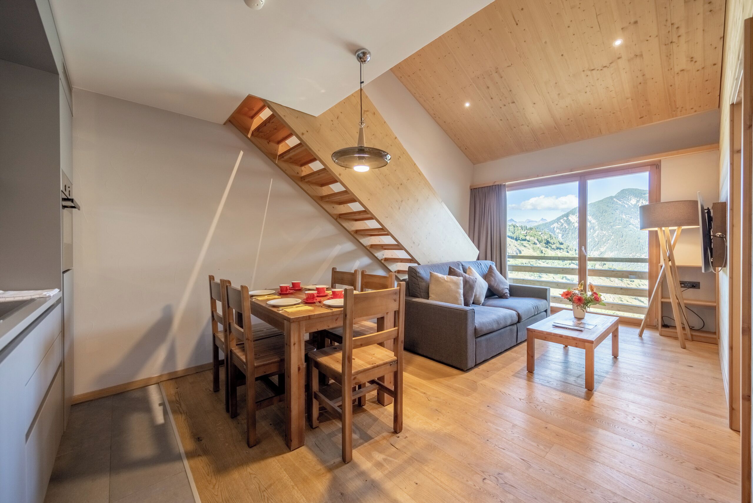 Quarto estilo loft com escada de madeira, mesa de jantar com cadeiras, sofá, mesa de centro e uma grande janela com vista para as montanhas.