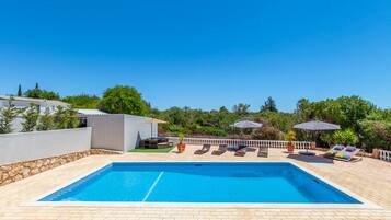 Uma piscina retangular com borda azulejada, rodeada por espreguiçadeiras e guarda-sóis.