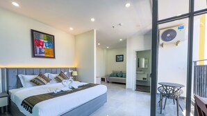 Quarto de hotel moderno com uma cama grande, um sofá, uma varanda com mesa e um banheiro visível através de uma porta aberta.