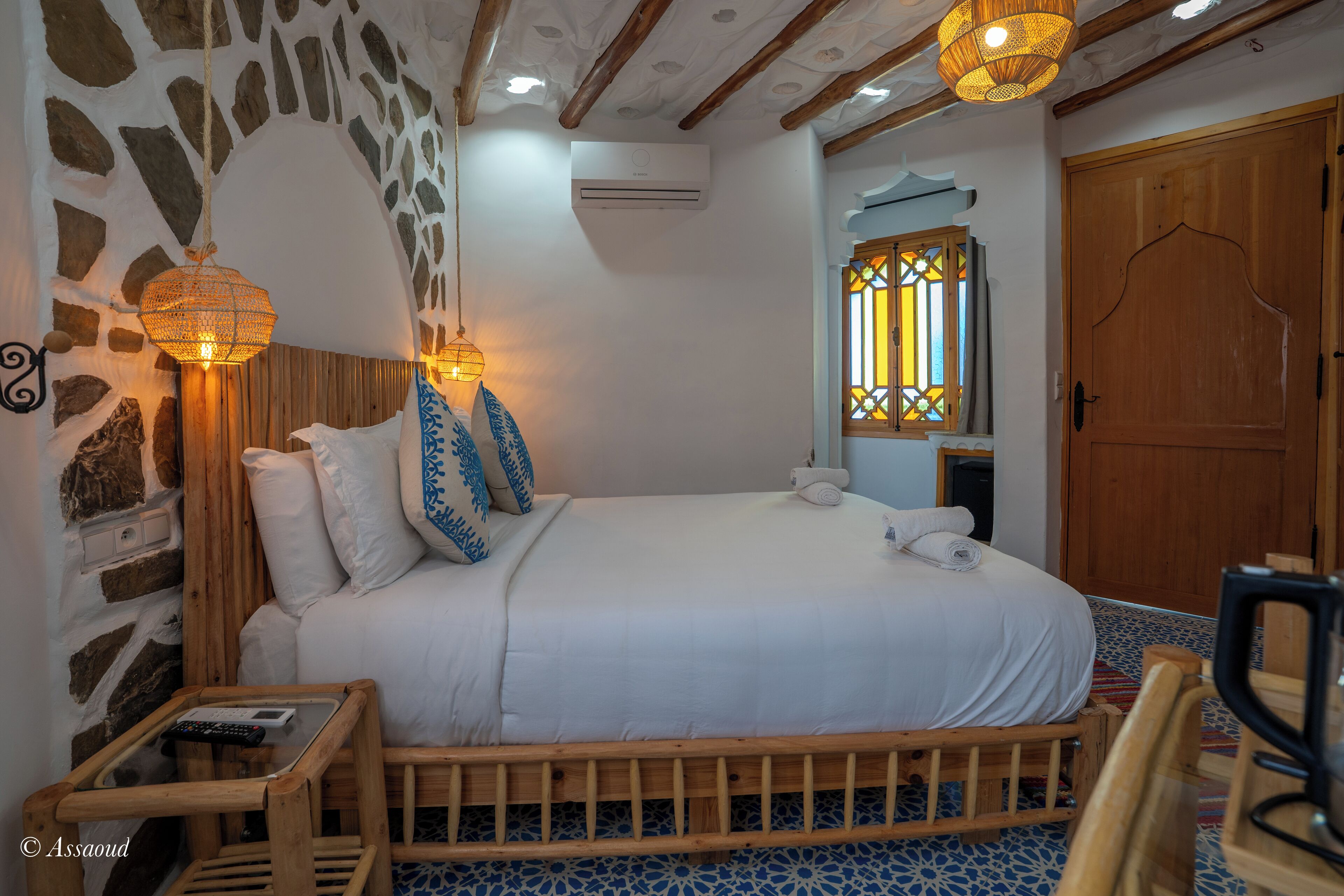 Quarto com parede de pedra, estrutura de cama de madeira, colcha branca e mesa de cabeceira de madeira com um controle remoto.