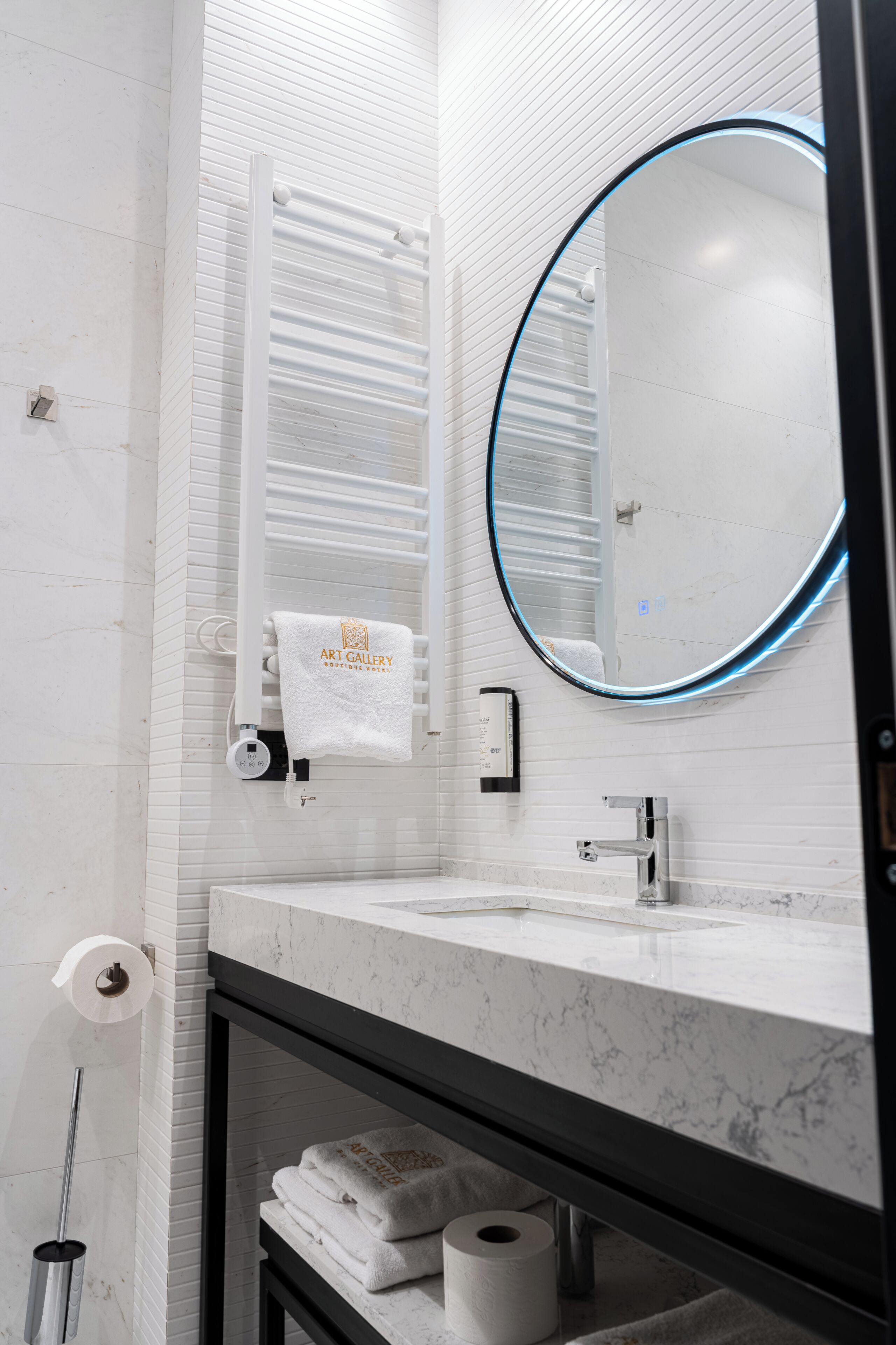 Um banheiro com parede em azulejos brancos, um espelho redondo com moldura azul, uma toalha branca pendurada na parede e uma prateleira preta com toalhas e um rolo de papel higiênico.