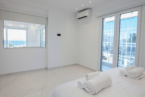 Quarto de hotel moderno com uma cama grande, vista da cidade através de janelas do chão ao teto, e uma varanda com vista para o oceano.