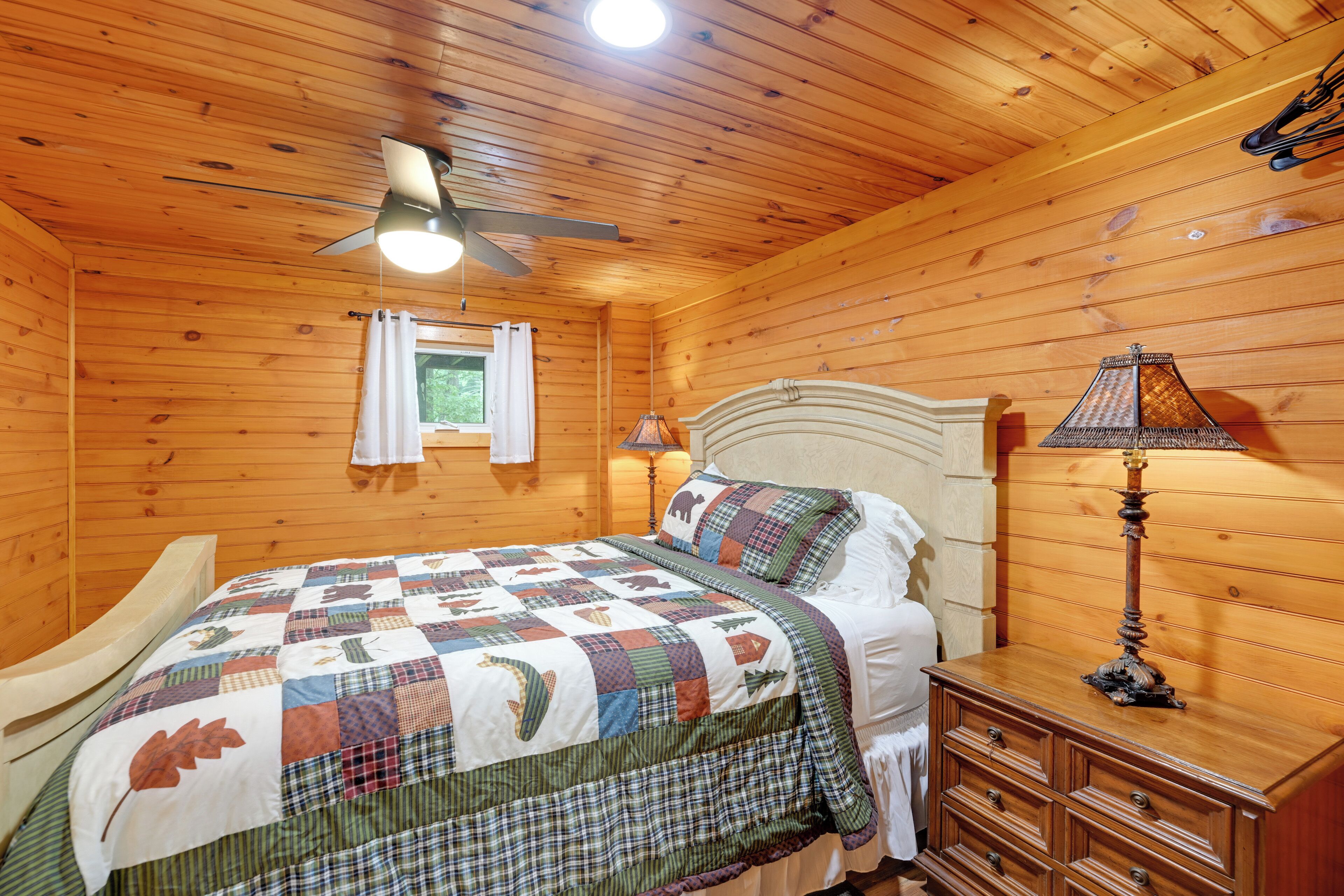 Quarto de cabana de madeira com cama, mesa de cabeceira, abajur e ventilador de teto.
