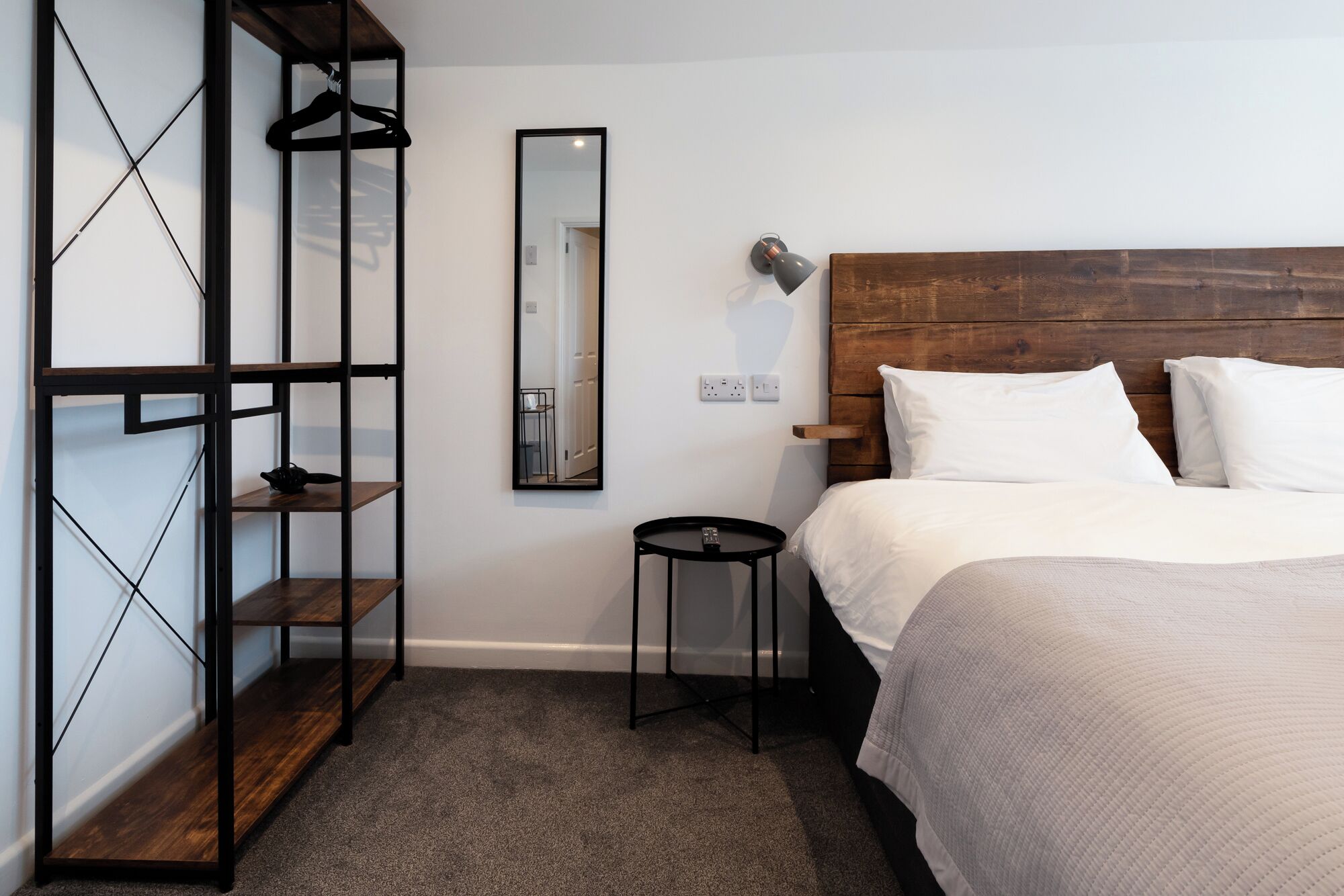 Quarto com cama, cabeceira de madeira, estante de metal preto, mesa redonda pequena e luminária fixada na parede.