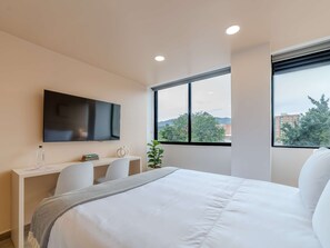 Quarto de hotel com cama, escrivaninha, cadeira, televisão e janela com vista para edifícios e árvores.