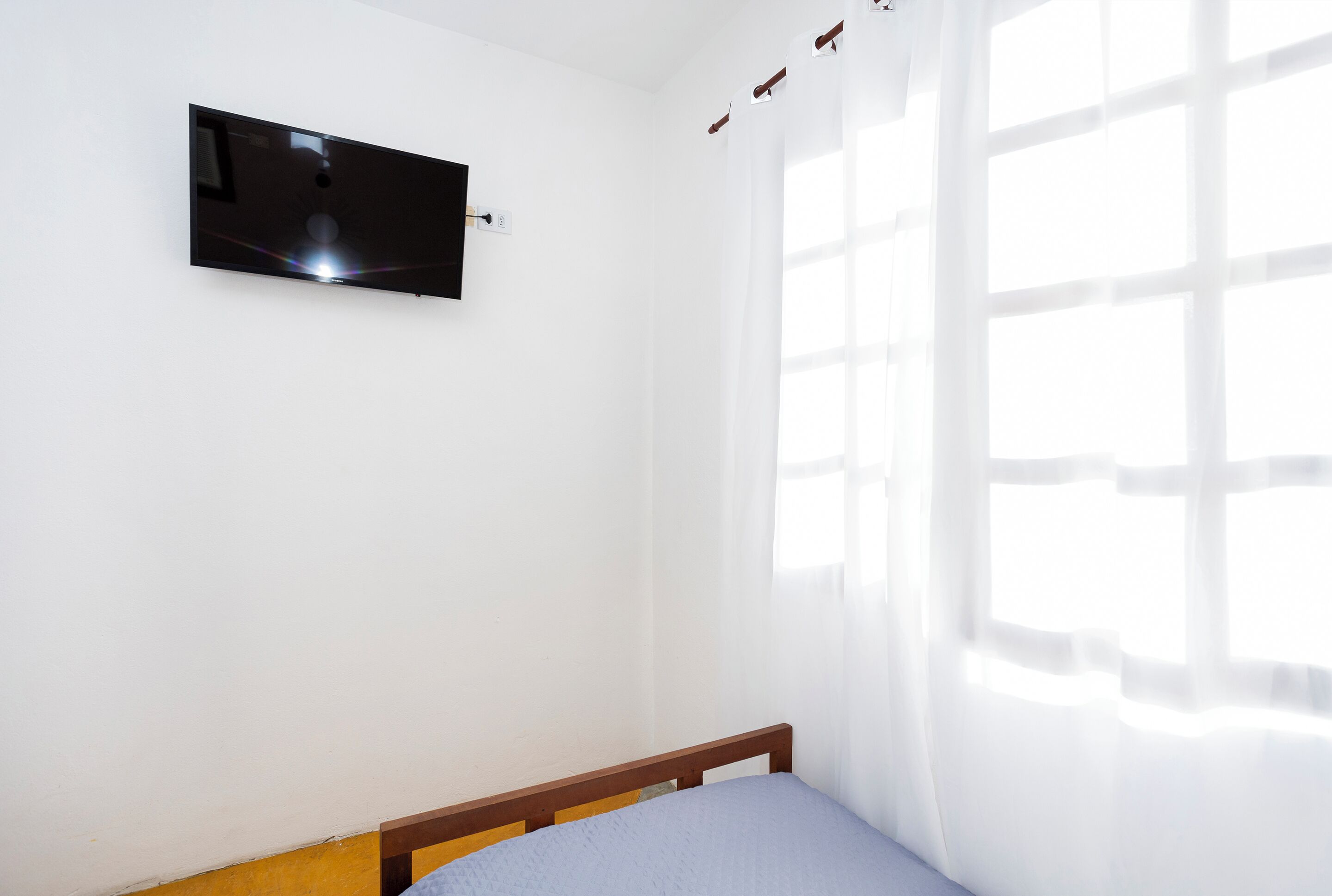 Quarto com cama, TV fixada na parede e janela com cortinas.