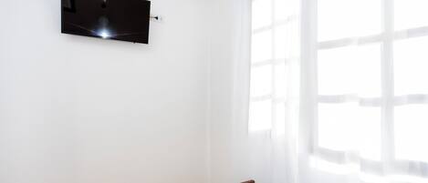 Quarto com cama, TV fixada na parede e janela com cortinas.