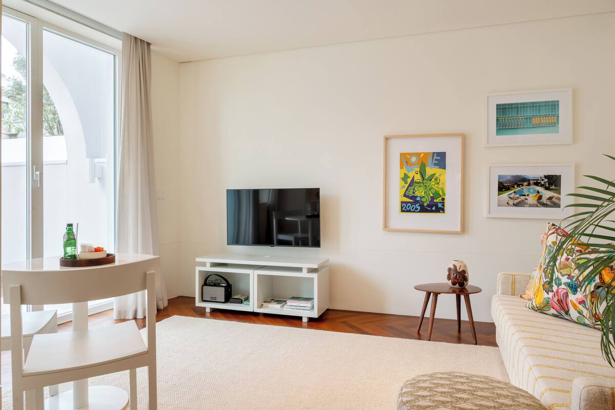 Uma sala de estar moderna com uma TV de tela plana sobre um suporte branco, uma mesinha lateral, um sofá e uma poltrona. Há quadros nas paredes e uma planta ao lado.
