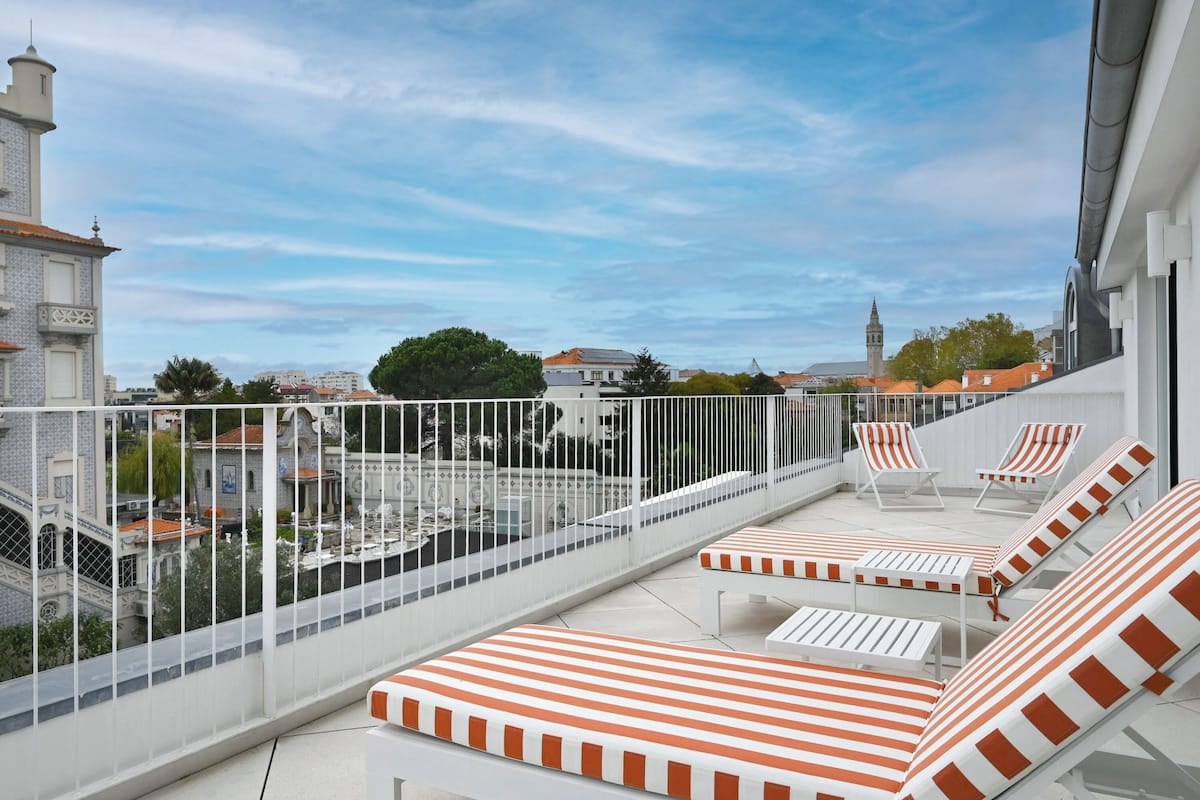 Um terraço no telhado com cadeiras de lounge listradas em branco e laranja, um corrimão branco e vista para prédios e uma torre ao fundo.