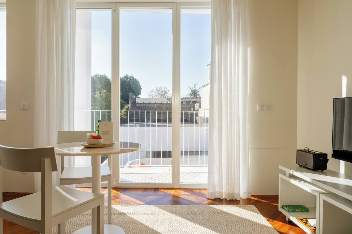 Quarto com uma mesa branca, cadeiras, uma televisão pequena e uma varanda com vista para prédios e árvores.