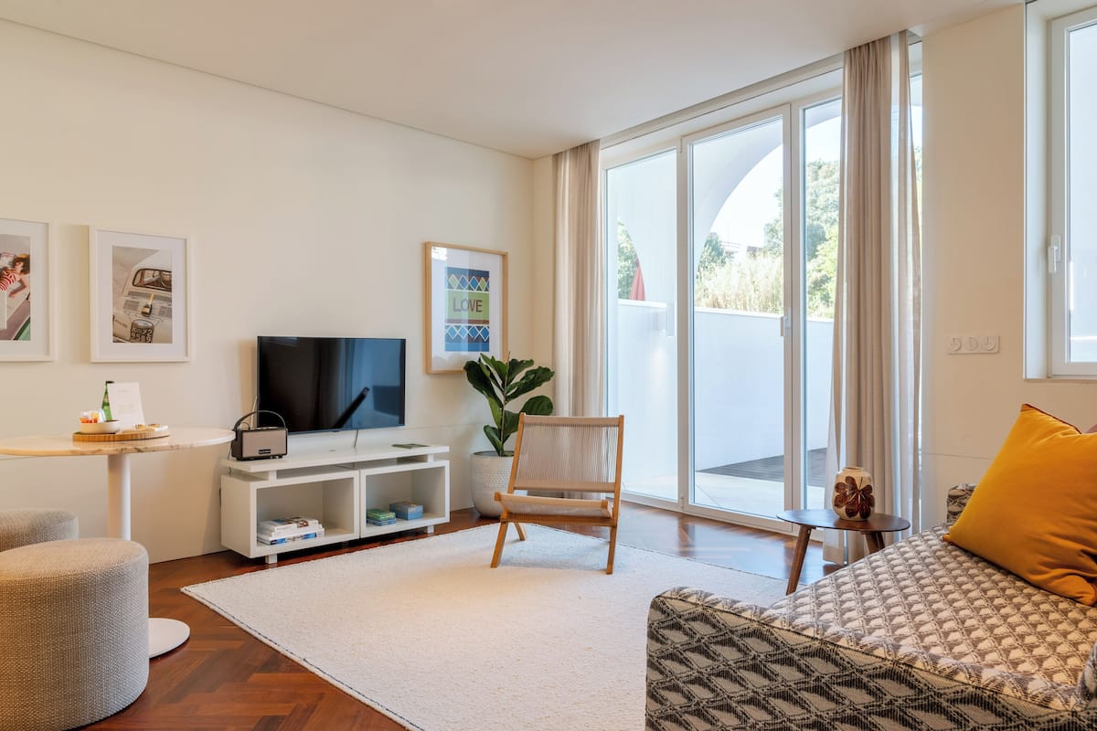 Uma sala de estar moderna com televisão de tela plana, um rack branco, uma janela grande com cortinas, um sofá estampado e uma pequena mesa lateral.