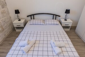 Cama bem arrumada com colcha estampada em padrão quadriculado, dois travesseiros e dois rolinhos de toalha sobre piso de madeira.