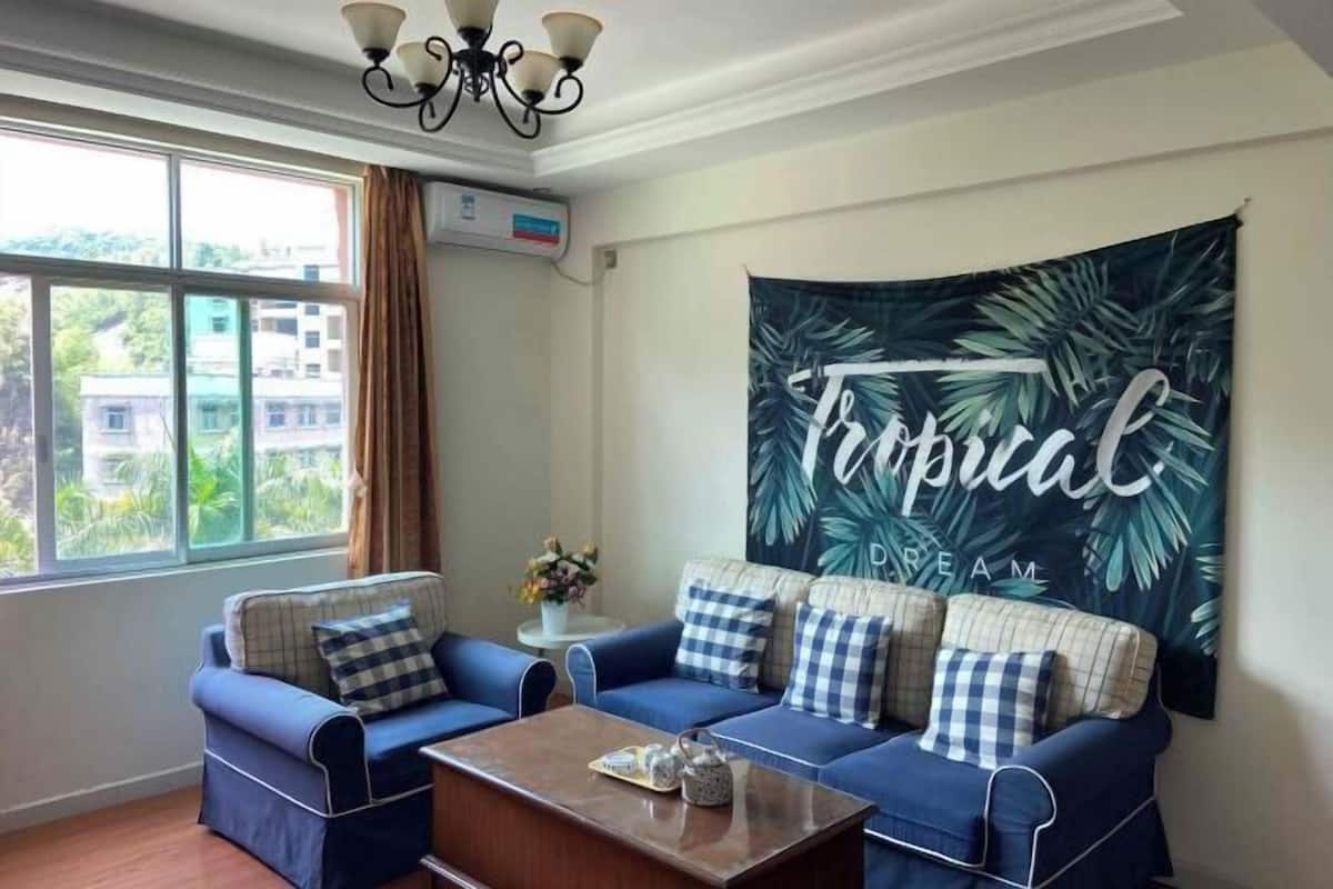 Quarto com sofá azul, mesa de centro de madeira e tapeçaria com tema tropical na parede.