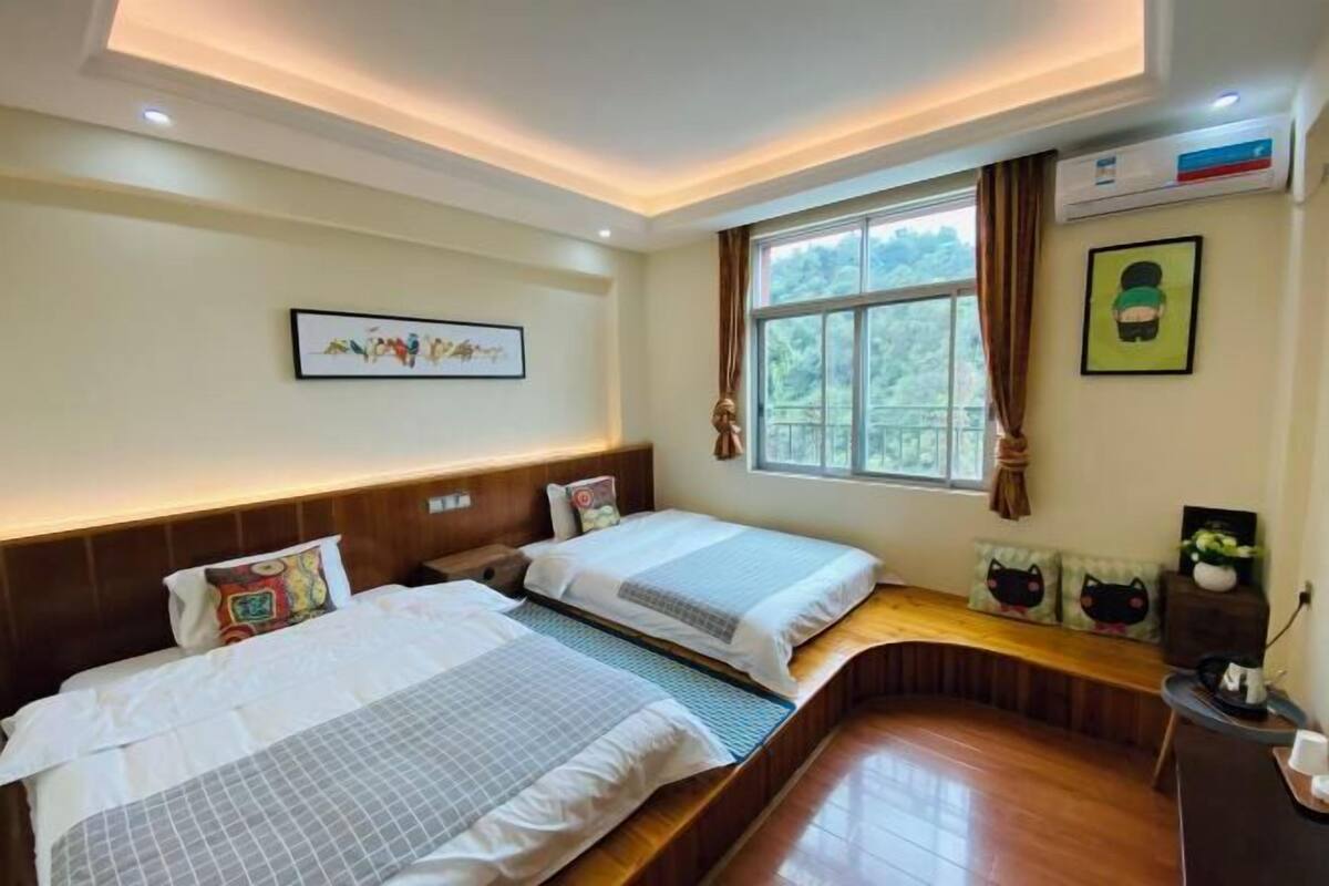 Quarto de hotel com duas camas, uma janela com cortinas, um banco de madeira e uma pequena mesa com uma planta.