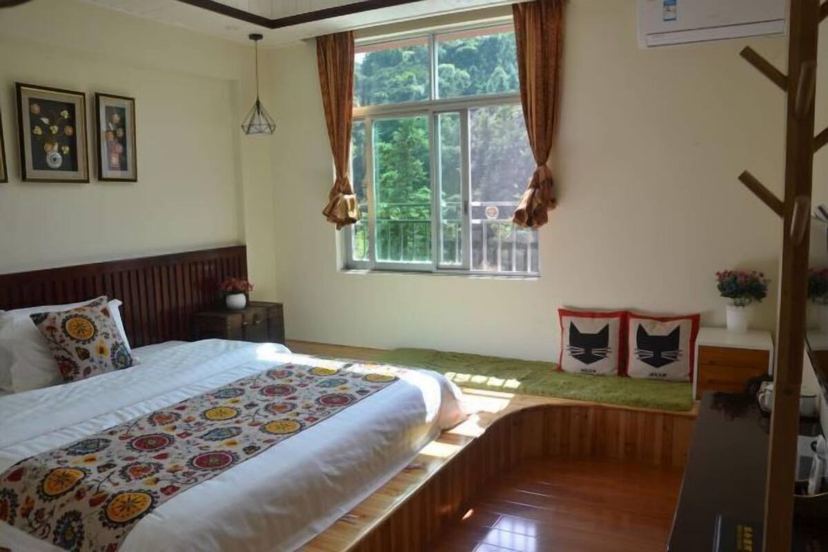 Quarto com uma cama grande, janela com cortinas, mesa de cabeceira com abajur e um banco com almofadas.
