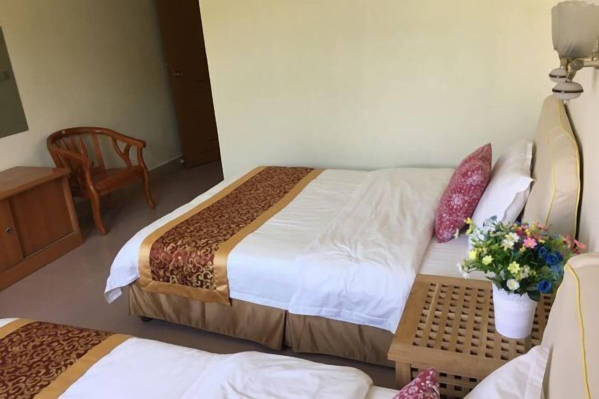Quarto de hotel com duas camas, uma cadeira de madeira, uma mesinha de cabeceira com abajur e um vaso de flores.