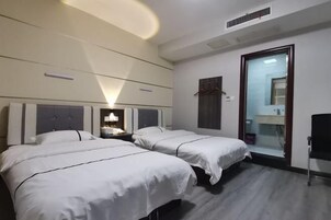 Quarto de hotel com duas camas, uma mesa de cabeceira com abajur, espelho e banheiro visível por uma porta aberta.