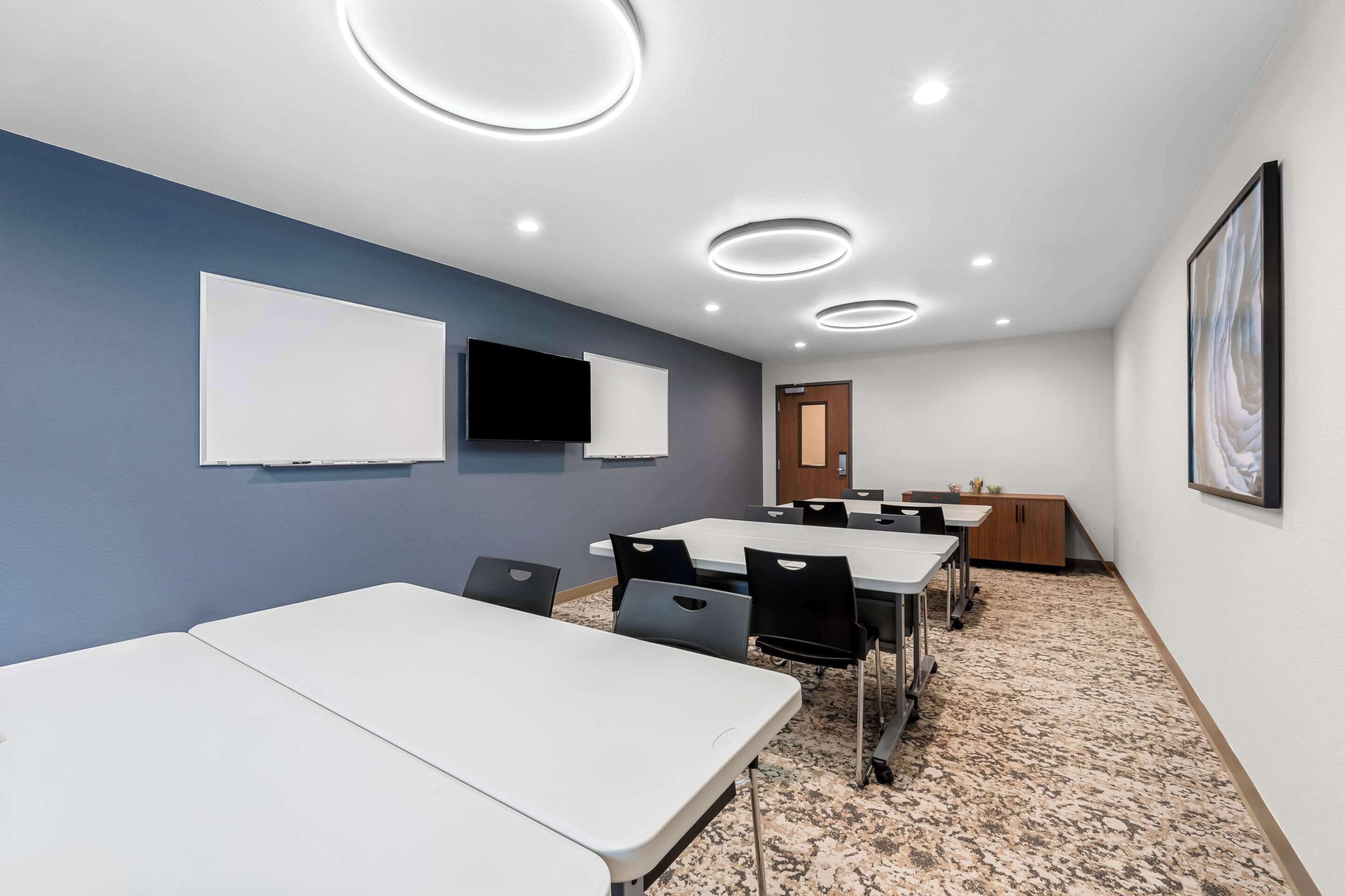 Uma sala de conferências com mesas e cadeiras brancas, uma TV de tela plana e um armário de madeira.