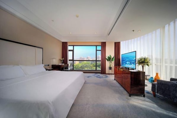 Quarto de hotel com uma cama grande, televisão, escrivaninha e vista da cidade através de amplas janelas.