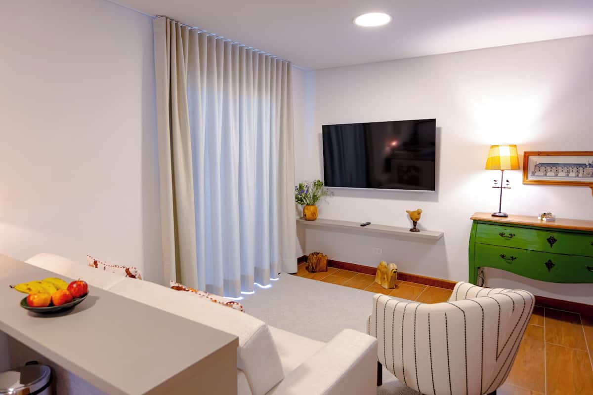 Quarto de hotel com uma cama, uma escrivaninha com uma cesta de frutas, uma cadeira, uma televisão, um abajur e uma cômoda verde.