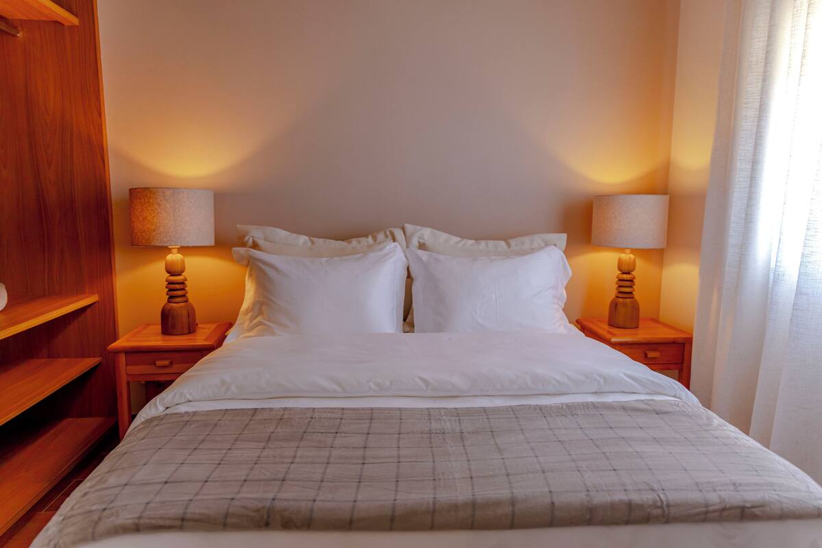 Uma cama bem arrumada com lençóis brancos e manta xadrez, com duas mesinhas de cabeceira de madeira e luminárias.