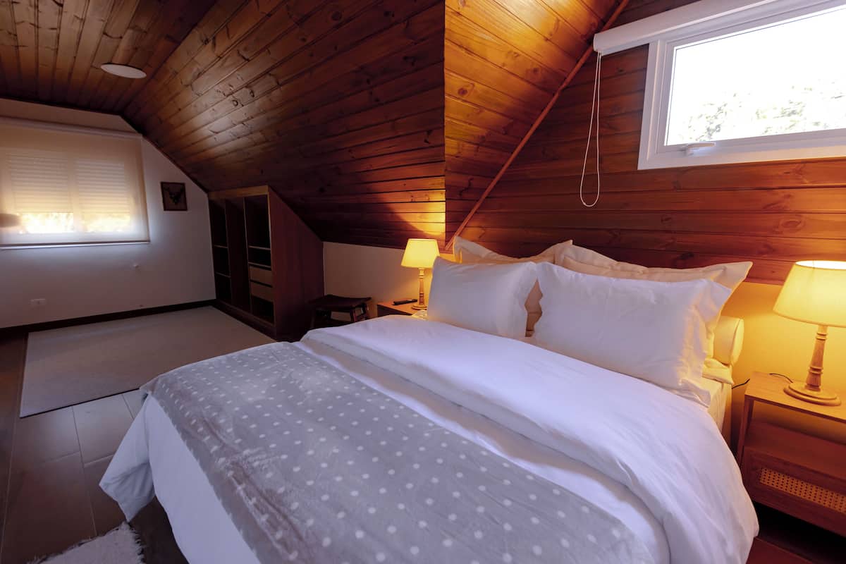 Quarto com teto de madeira, cama com roupa de cama branca, duas luminárias de cabeceira e uma janela com persianas.