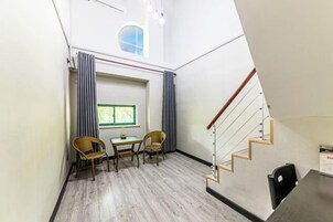 Quarto pequeno e moderno com piso de madeira, uma mesa, duas cadeiras, uma janela com cortinas e uma escada com corrimão de madeira.