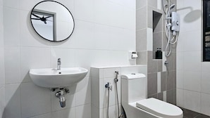 Casa de banho moderna com lavatório branco, sanita e chuveiro. Um espelho redondo está pendurado na parede, e é visível um caixote do lixo azul.
