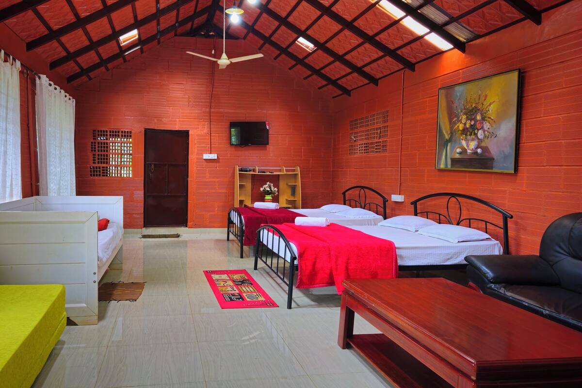 Quarto com teto alto, vigas de madeira, cama com cobertor vermelho, sofá amarelo, mesa de madeira e um quadro na parede.