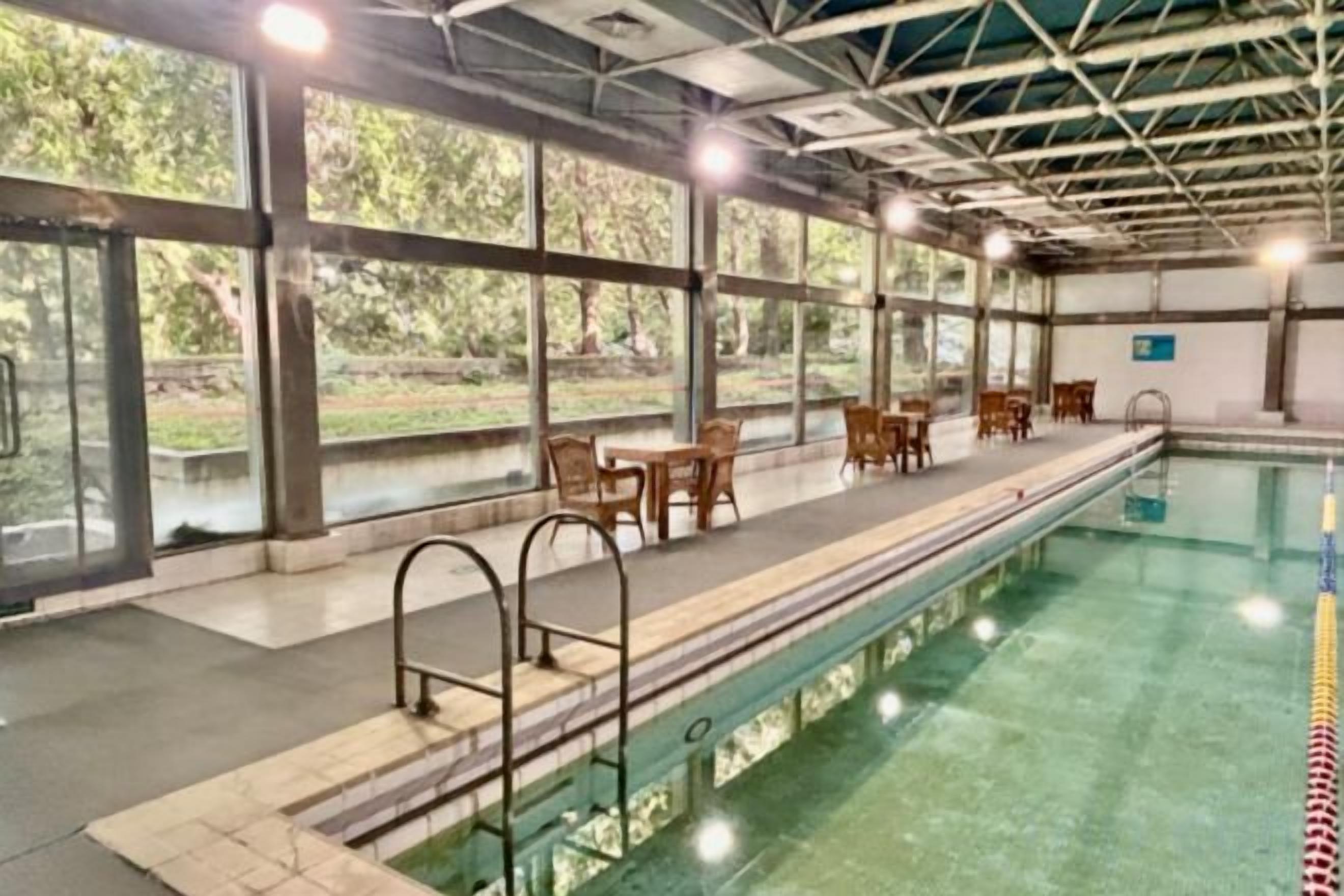Piscina interna com paredes de vidro transparente, assentos de deck em madeira e escada de metal.