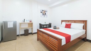 Quarto de hotel com uma cama, cabeceira de madeira, almofada vermelha, pequena mesa com uma planta, prateleira com objetos e um refrigerador.