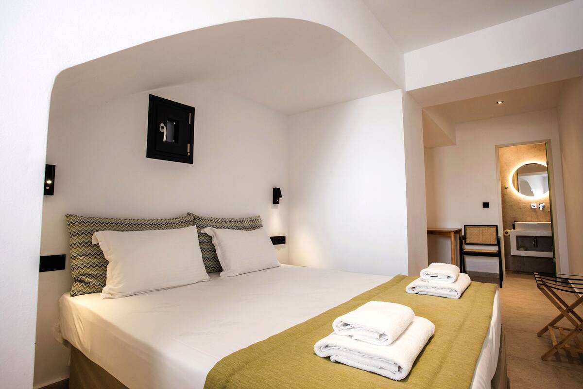 Quarto de hotel com cama bem arrumada, banheiro pequeno com espelho e porta-toalhas.