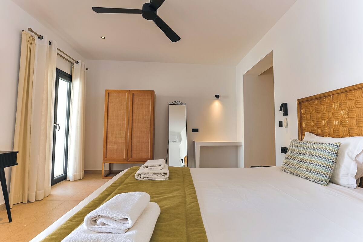 Quarto de hotel com cama, ventilador de teto, guarda-roupa e banheiro visível por uma porta aberta.